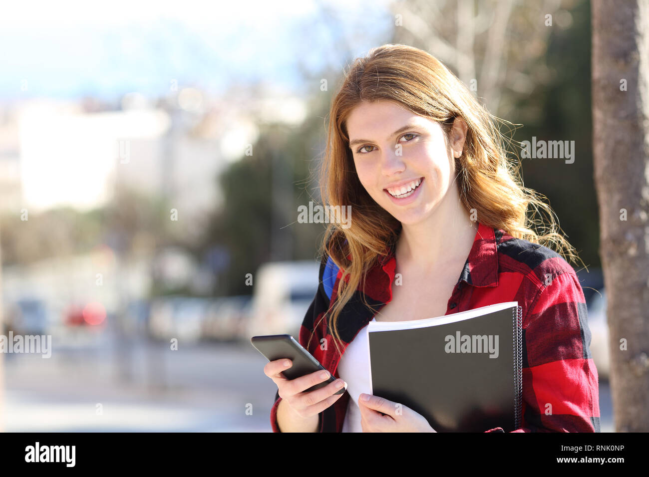 Happy student holding smart phone and notebook looking at camera in the ...
