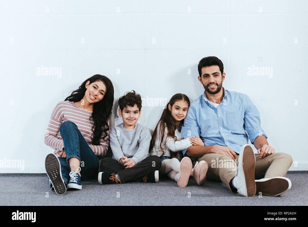 cheerful hispanic family smiling while sitting on floor near white wall ...