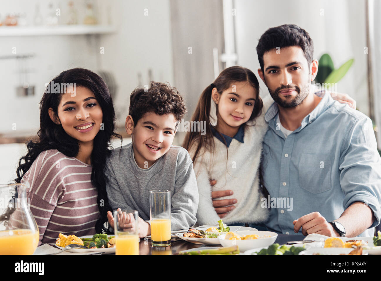 cheerful latin family hugging and looking at camera during lunch at ...