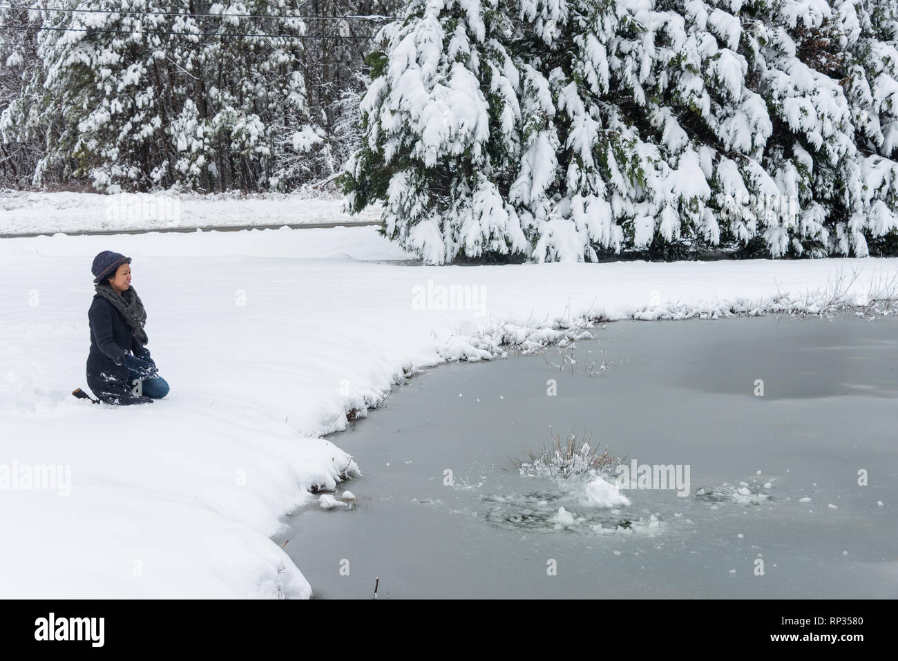 Asian woman with hat and winter coat kneeling in front of frozen pond ...