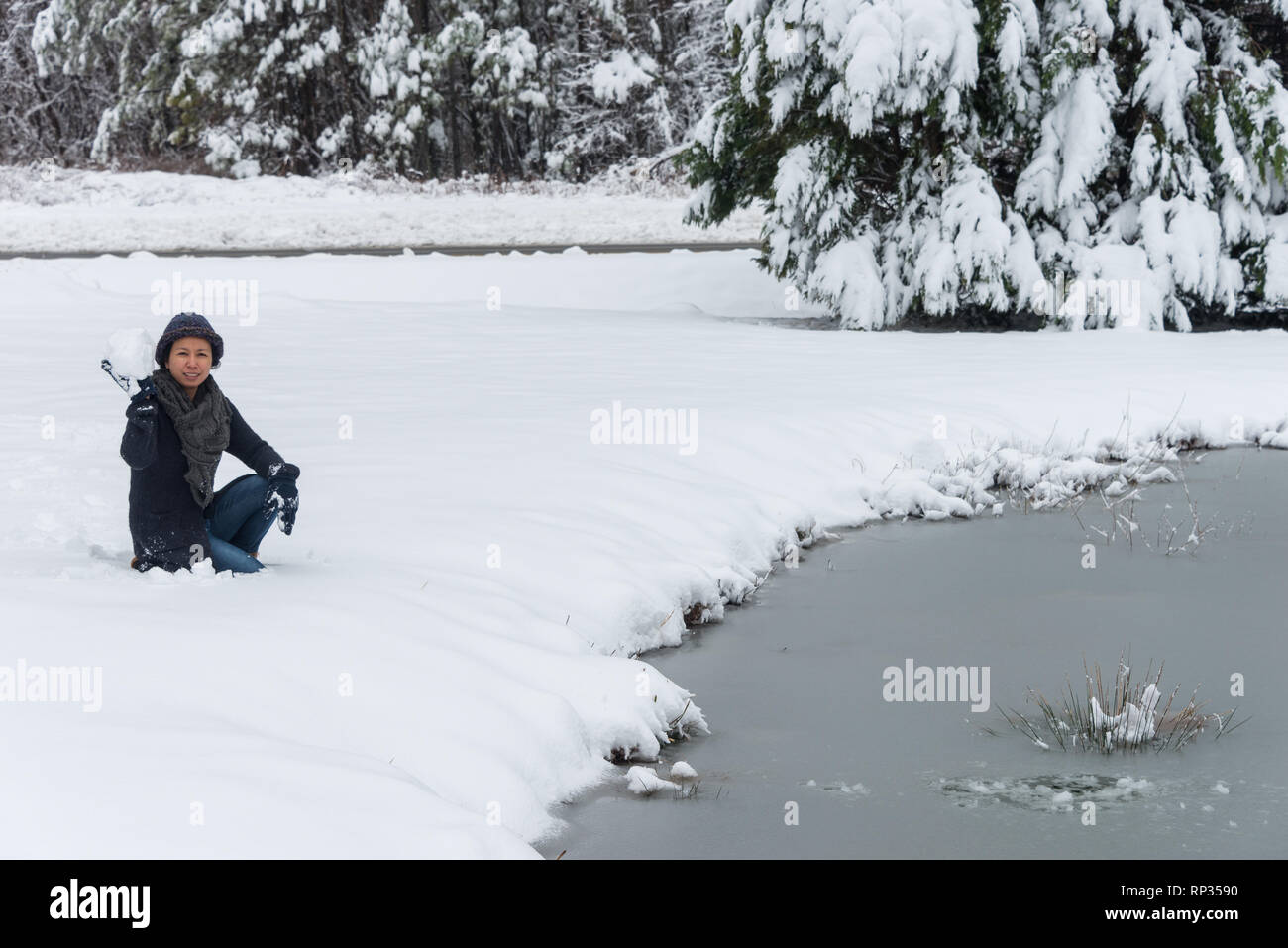 Asian woman with hat and winter coat kneeling in front of frozen pond ...
