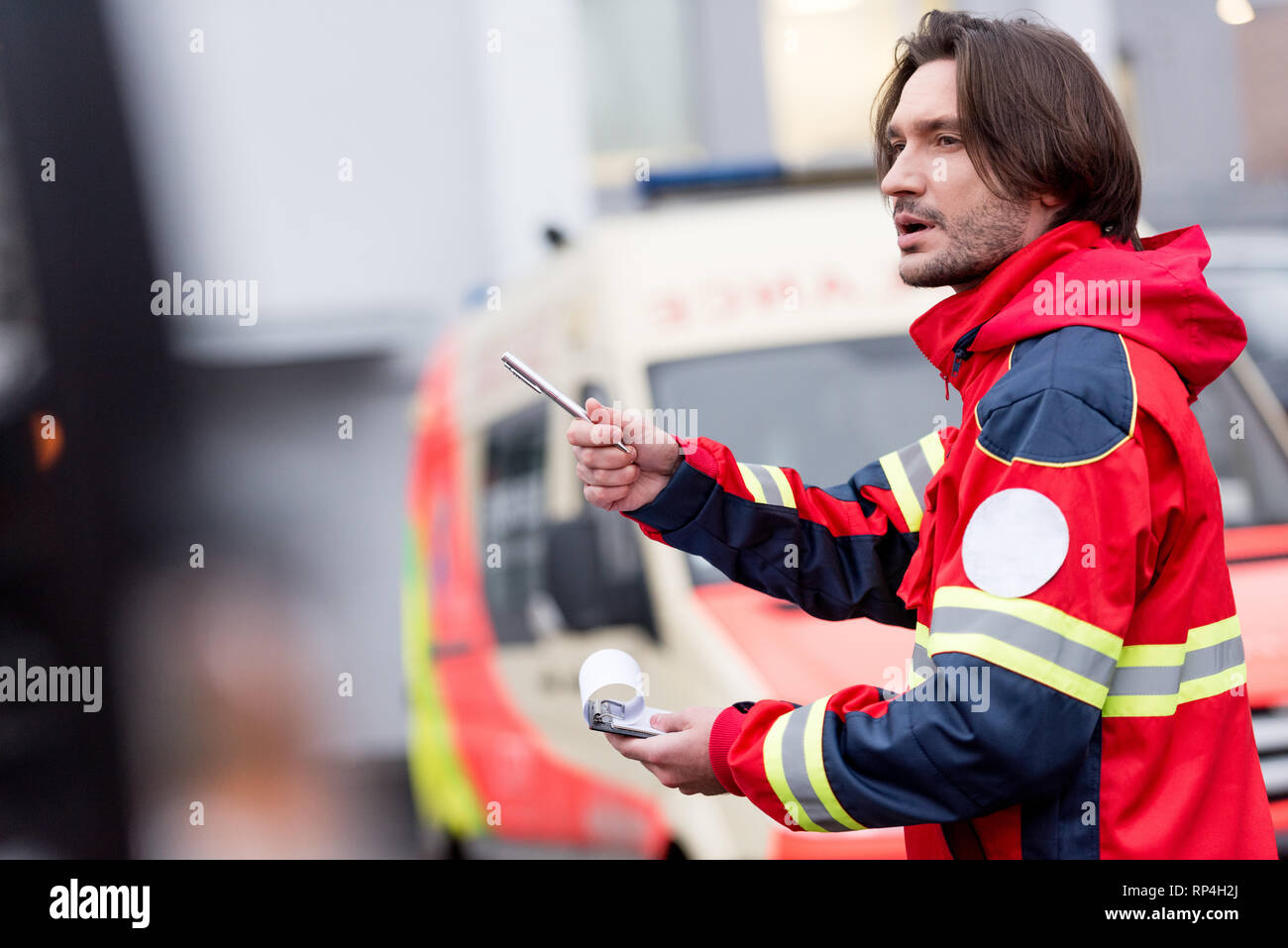 Brunette paramedic in red uniform holding pen and clipboard on street ...