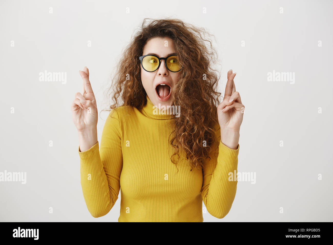 Excited woman in yellow sweater keeping fingers crossed, mouth wide ...