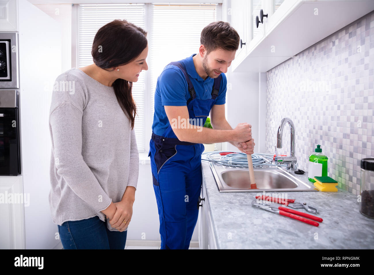 Happy Woman Looking At Male Plumber Using Plunger In The Kitchen Sink ...