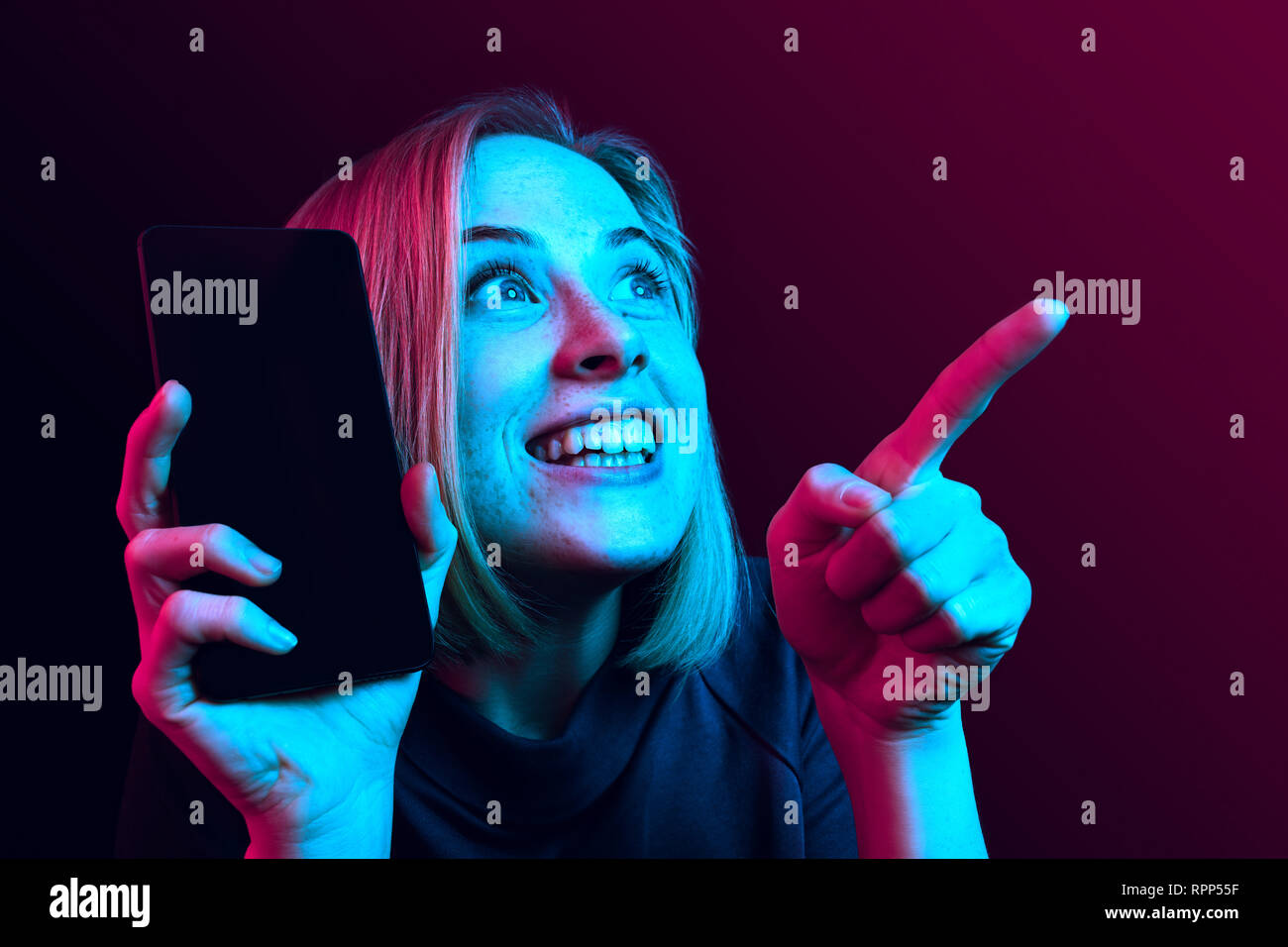 Portrait of a confident casual girl showing blank screen of mobile phone over neon studio ...