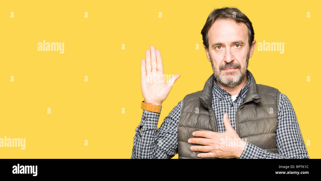 Middle age handsome man wearing winter vest Swearing with hand on chest ...