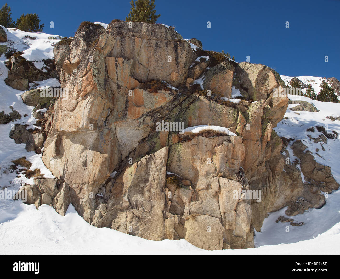 A cliff on a snowy slope against the blue sky with a lone tree on top ...