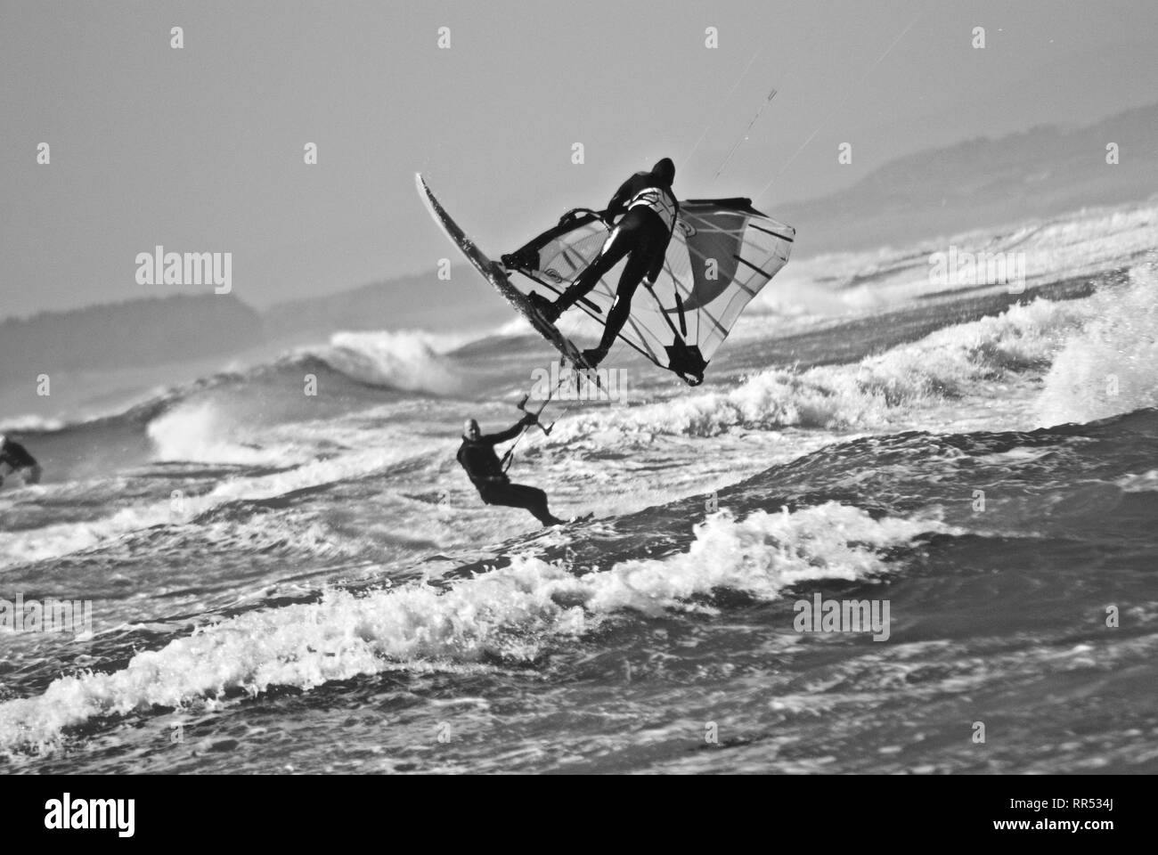 A wind surfer becomes airborne on dramatic white water in grey overcast ...