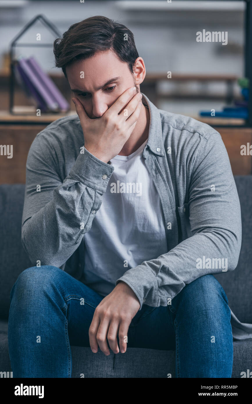 depressed man sitting, covering mouth with hand and grieving at home ...
