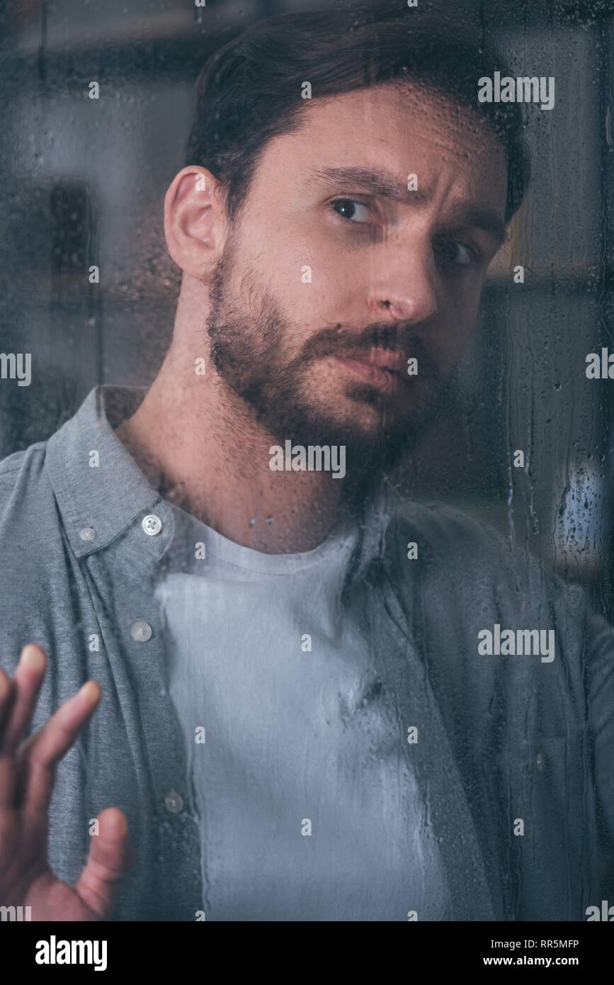 handsome upset man touching window with raindrops and looking away ...