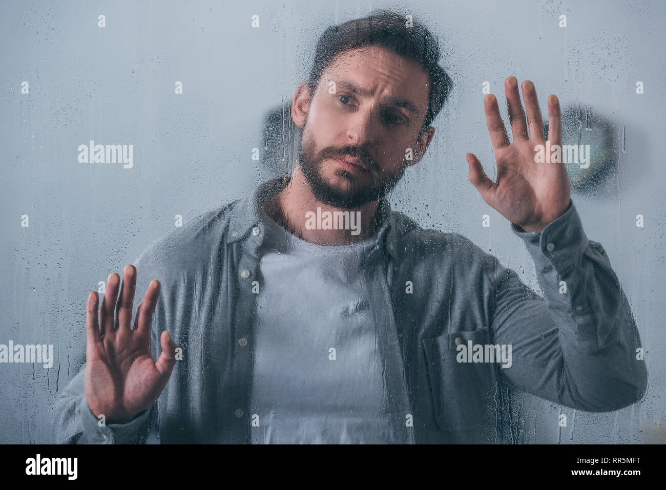 handsome upset man touching window with raindrops and looking away ...
