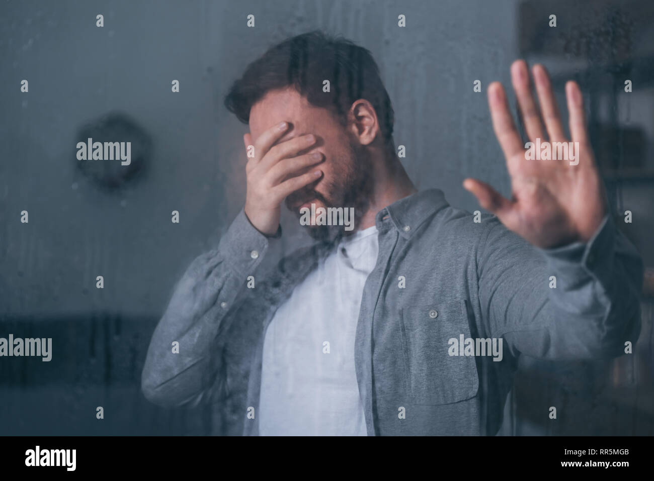 upset man covering face with hand, crying and touching window with ...