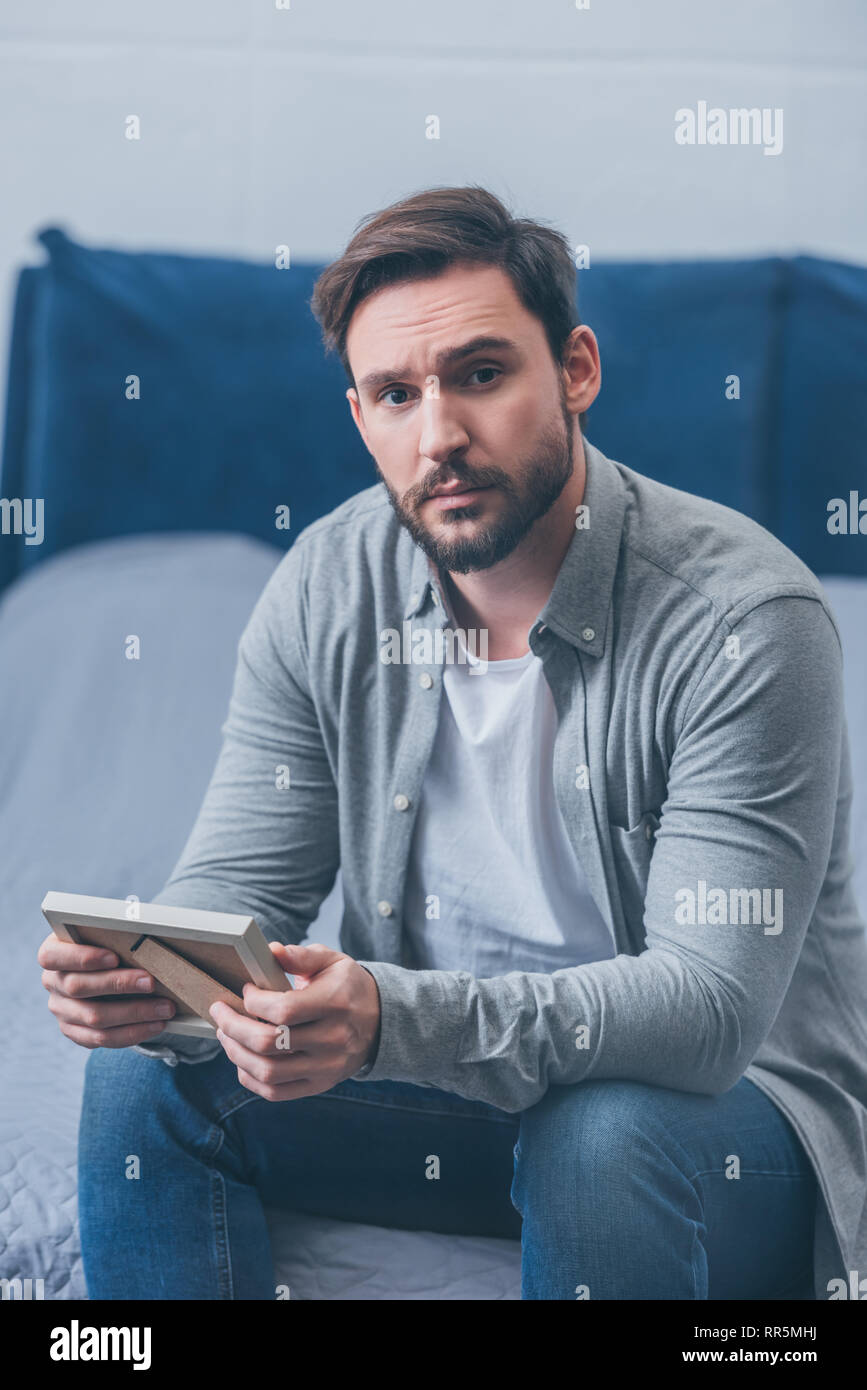 sad man sitting on bed, looking at camera and holding photo frame at ...