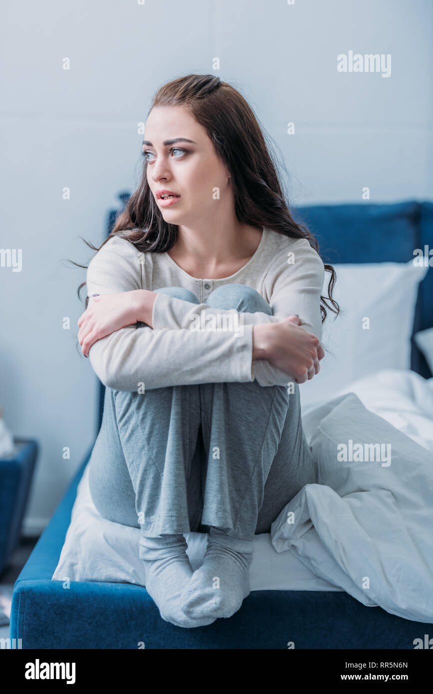 upset woman hugging knees, sitting on bed at home and looking away ...