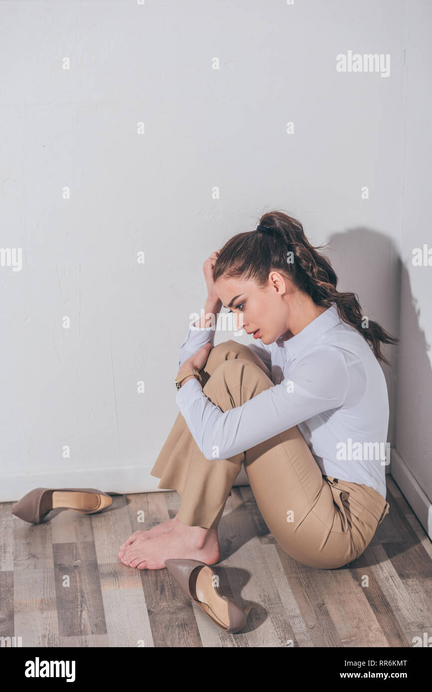 sad woman in white blouse and beige pants sitting on floor near wall at ...