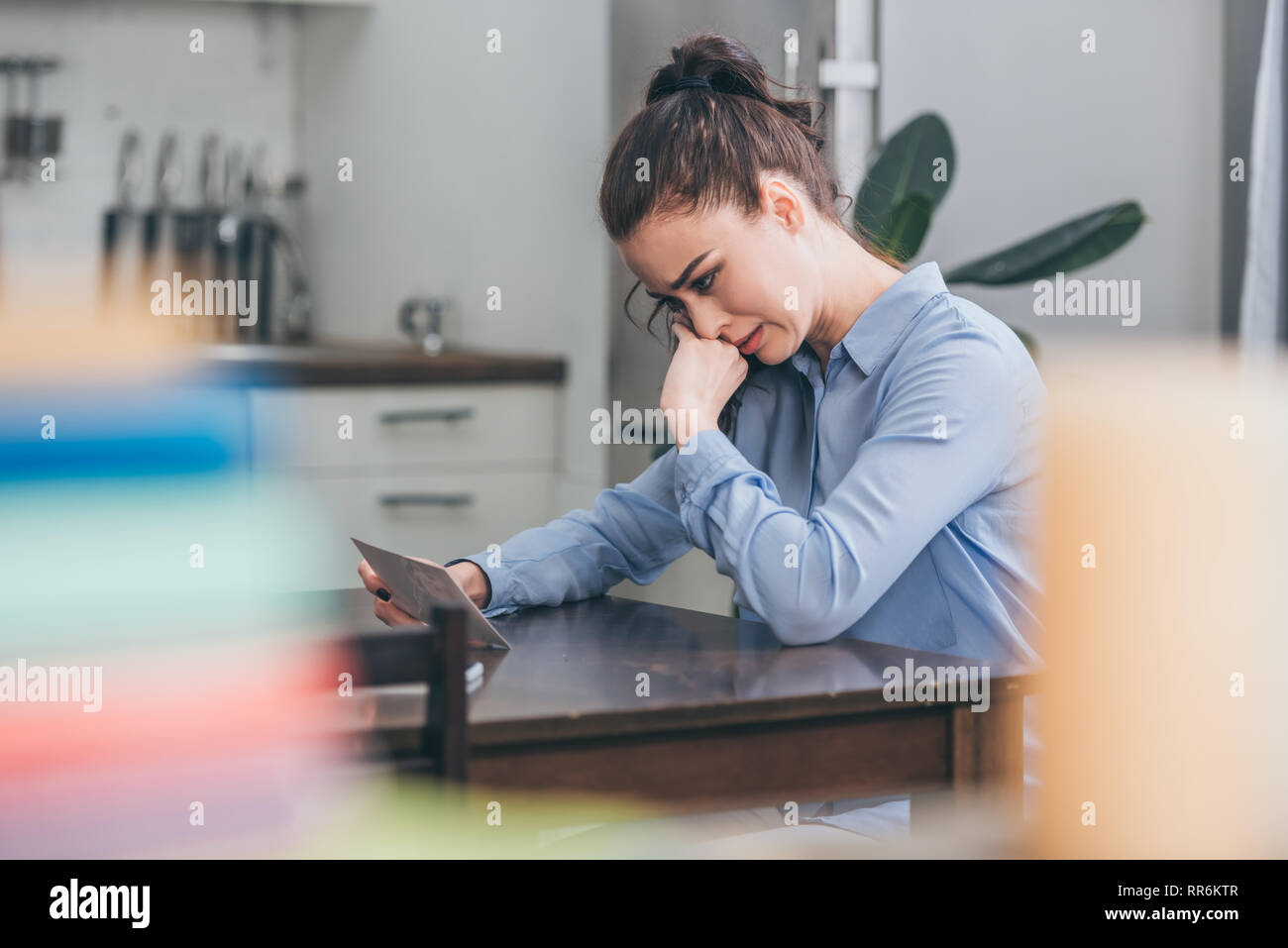 sad woman in blue blouse sitting at wooden table, looking at photo and ...