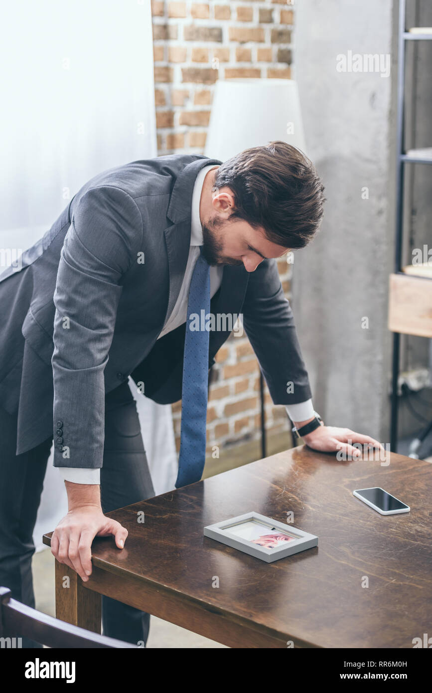 upset man in gray suit standing near table with smartphone and looking ...