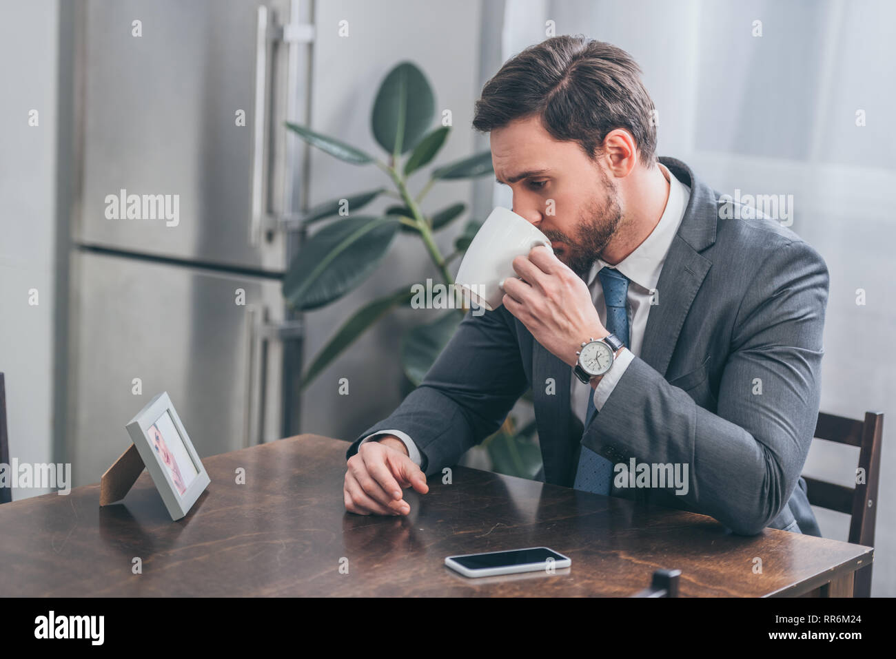 sad man in gray suit sitting at wooden table with smartphone drinking ...