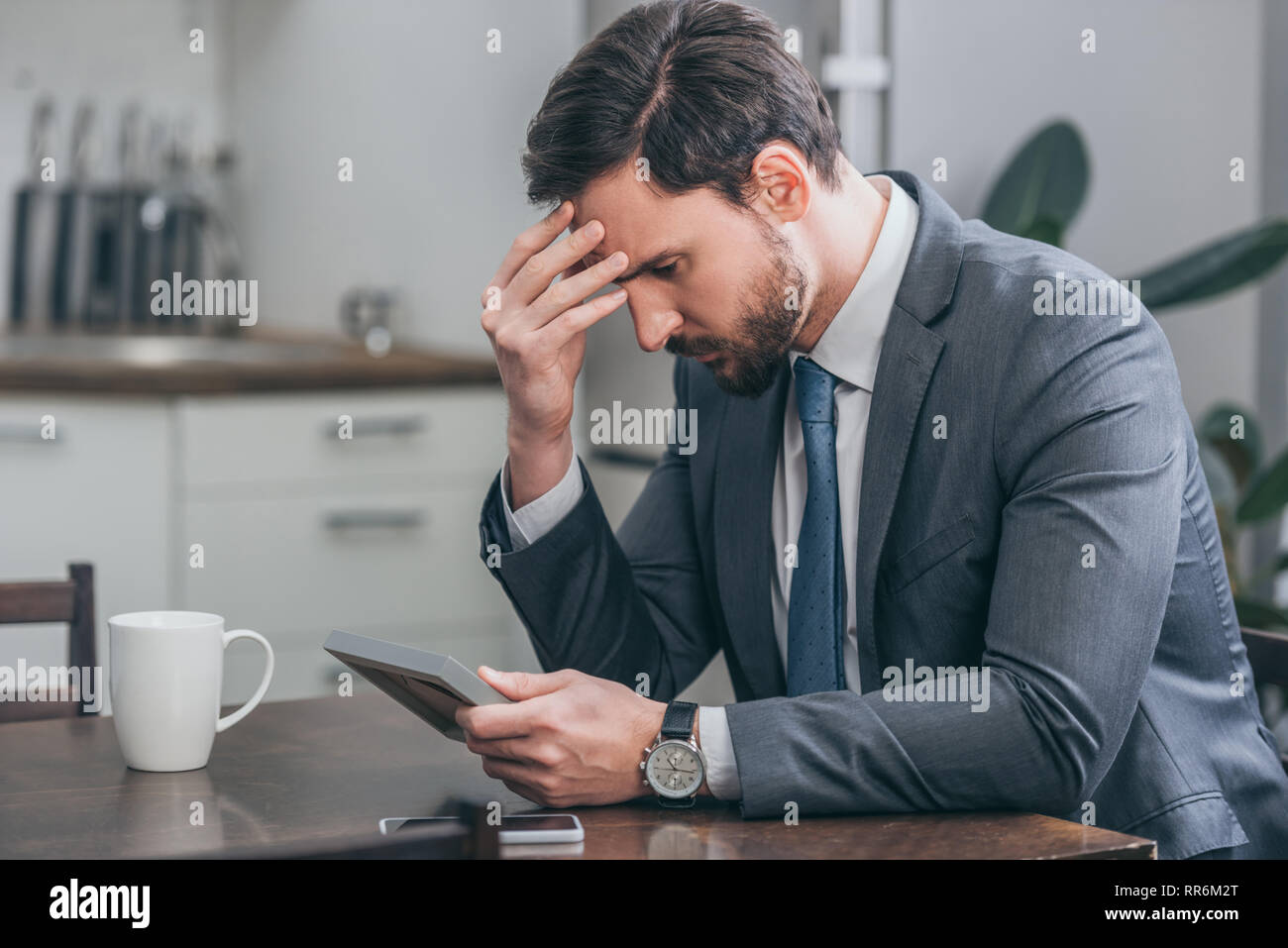sad man in gray suit sitting at wooden table with smartphone, white cup ...