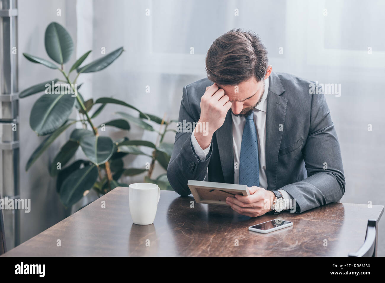 sad man in gray suit sitting at wooden table with smartphone, white cup ...