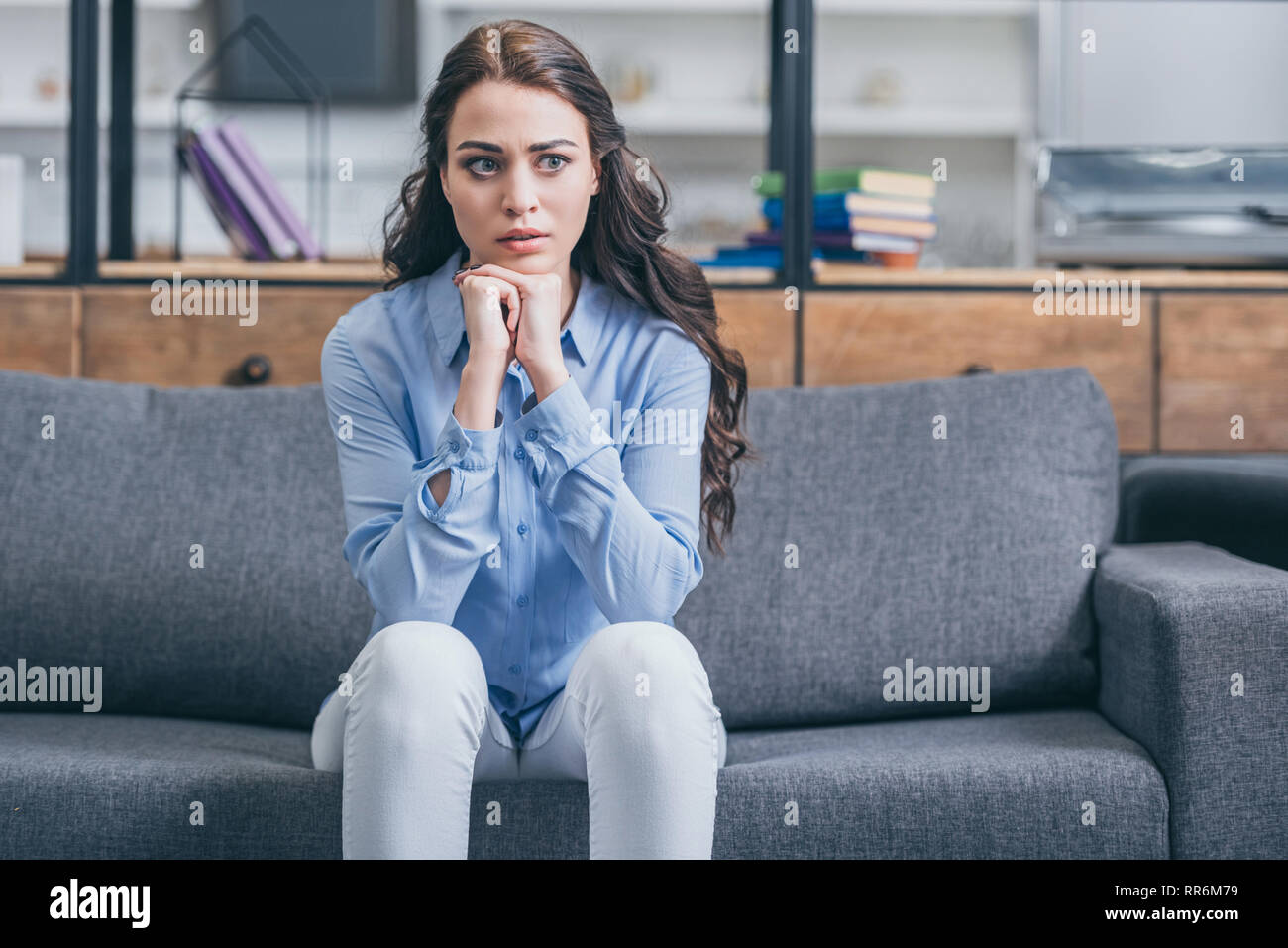 sad woman in blue blouse and white pants sitting on grey couch in ...