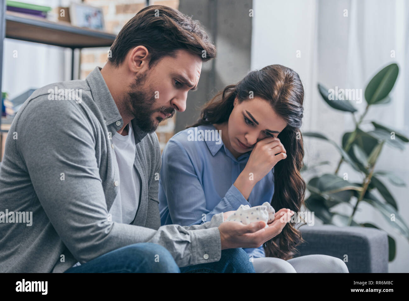 woman sitting and crying on couch while man holding baby shoes in room ...