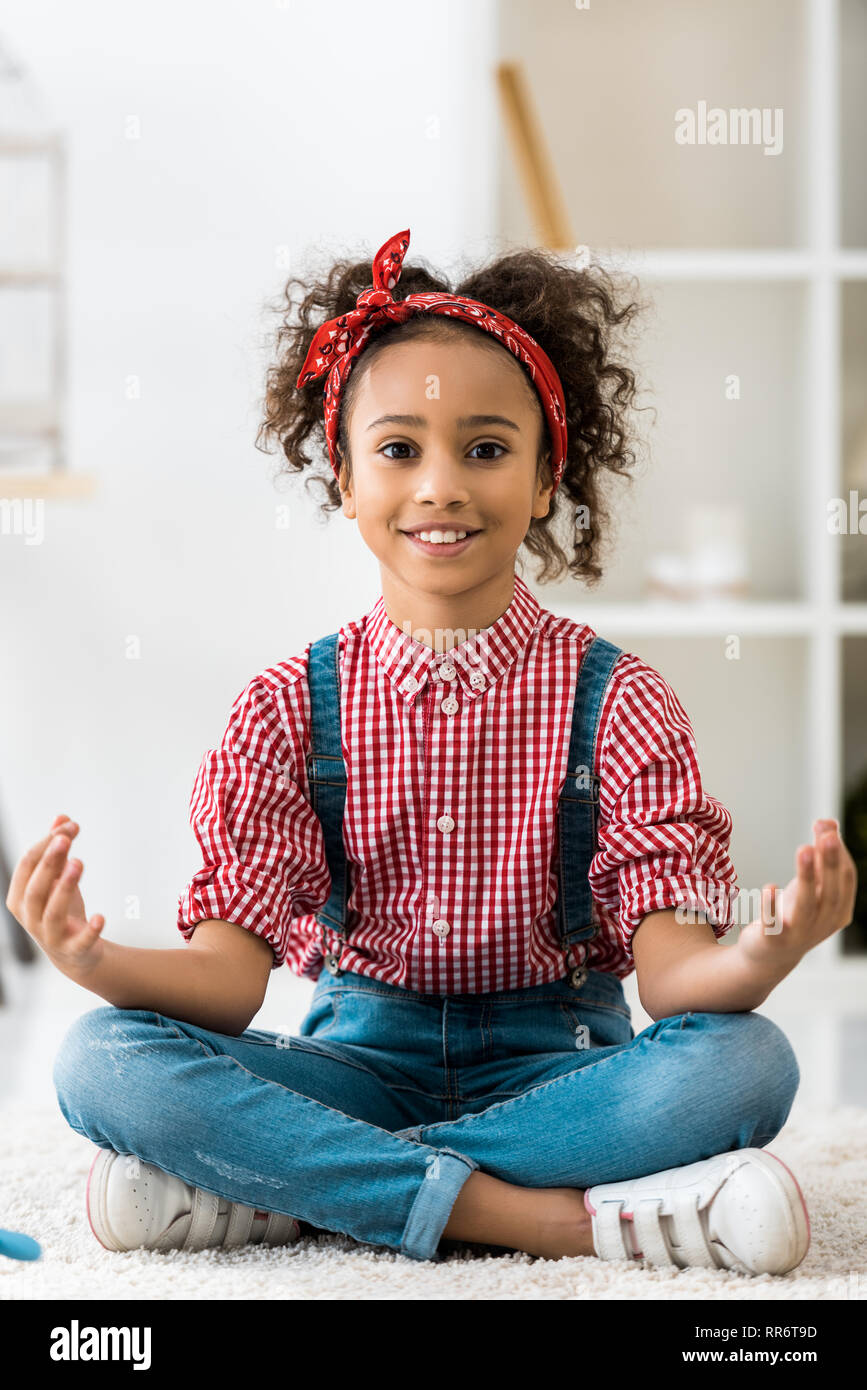 smiling african american child sitting in lotus pose and looking at ...