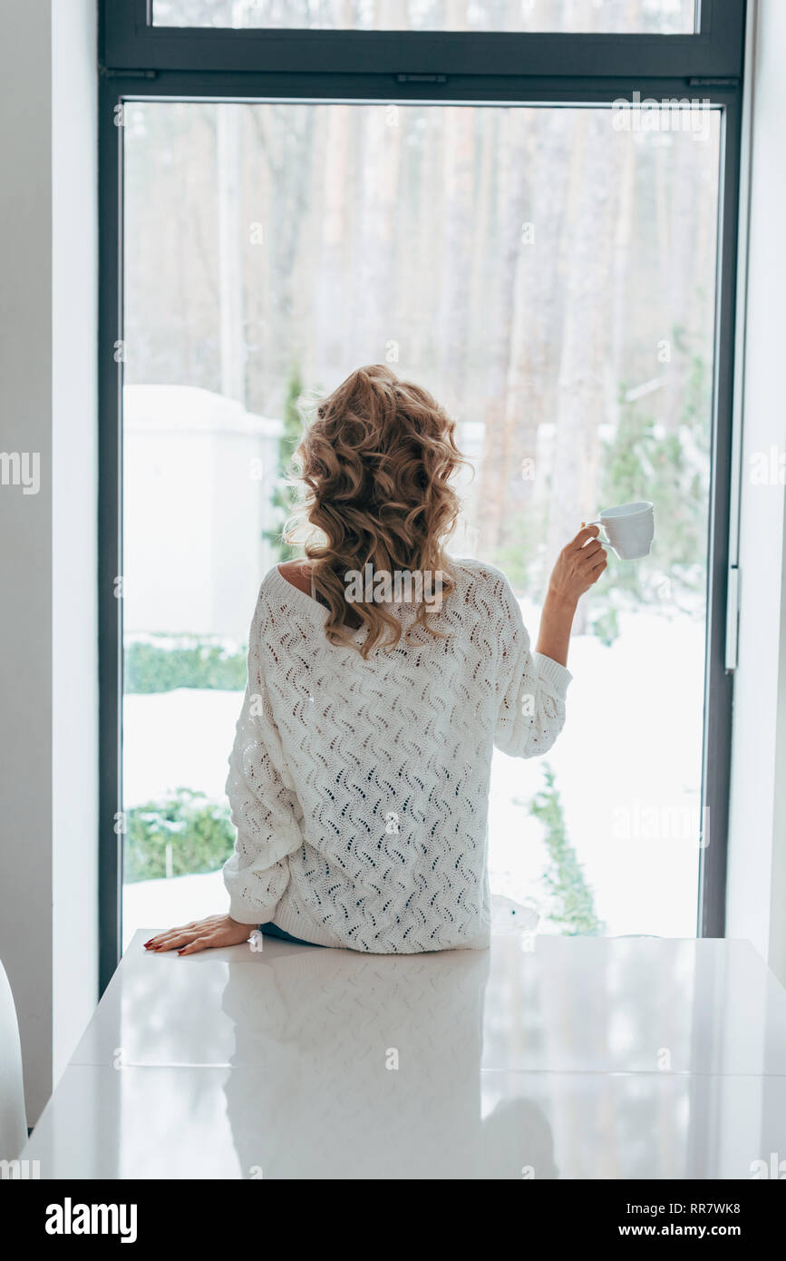 Back view of curly girl standing in front of window with coffee cup ...