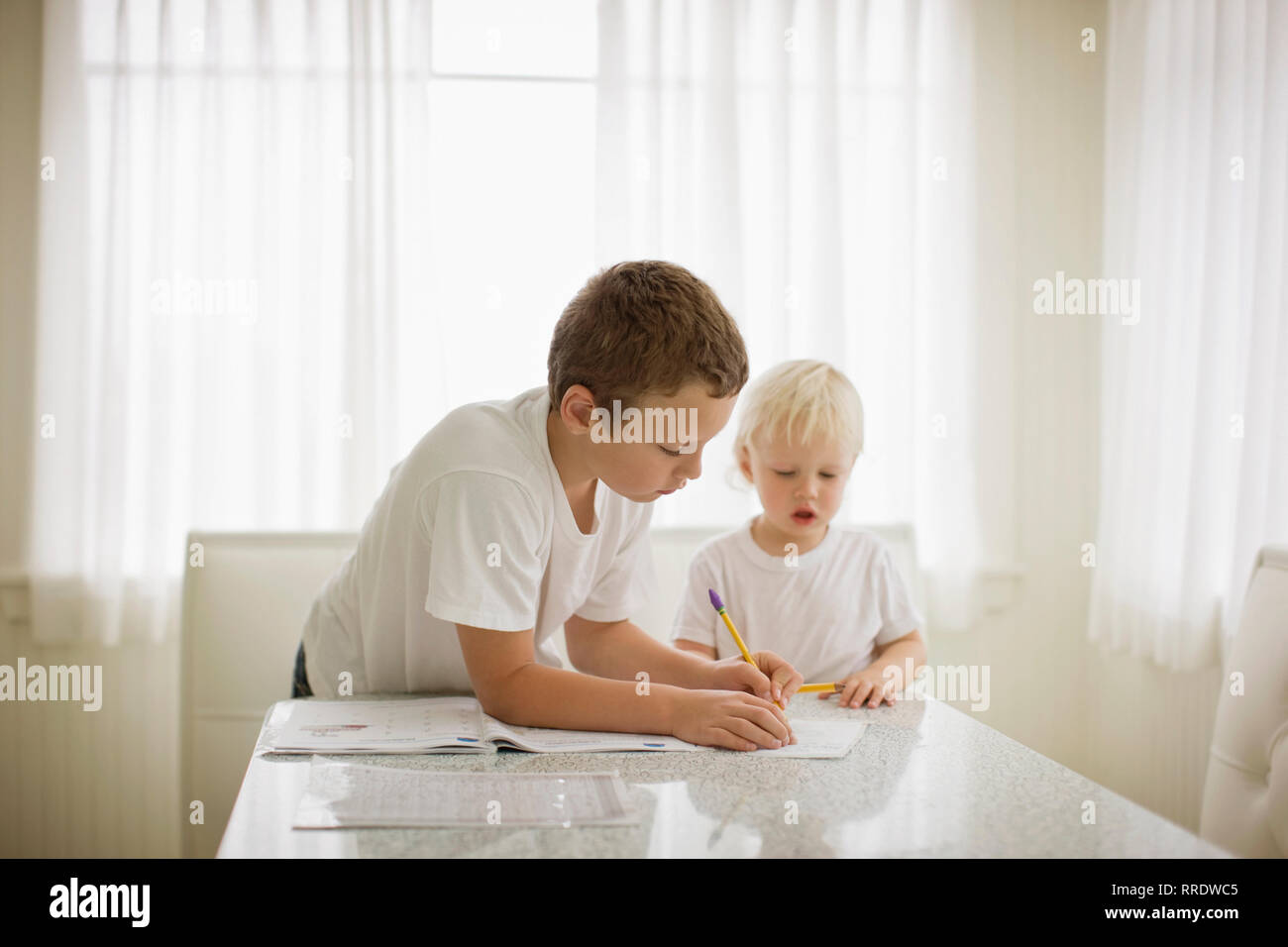 Young boy sitting at a dining table doing his homework with his younger ...