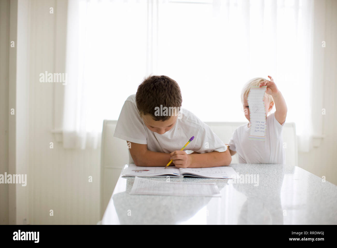 Young boy sitting at a dining table doing his homework with his younger ...