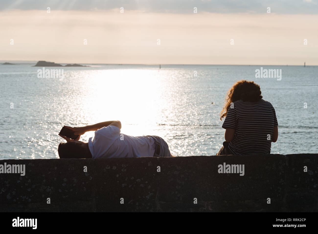 Rear view of couple fooling around while sitting on wall during sunset ...