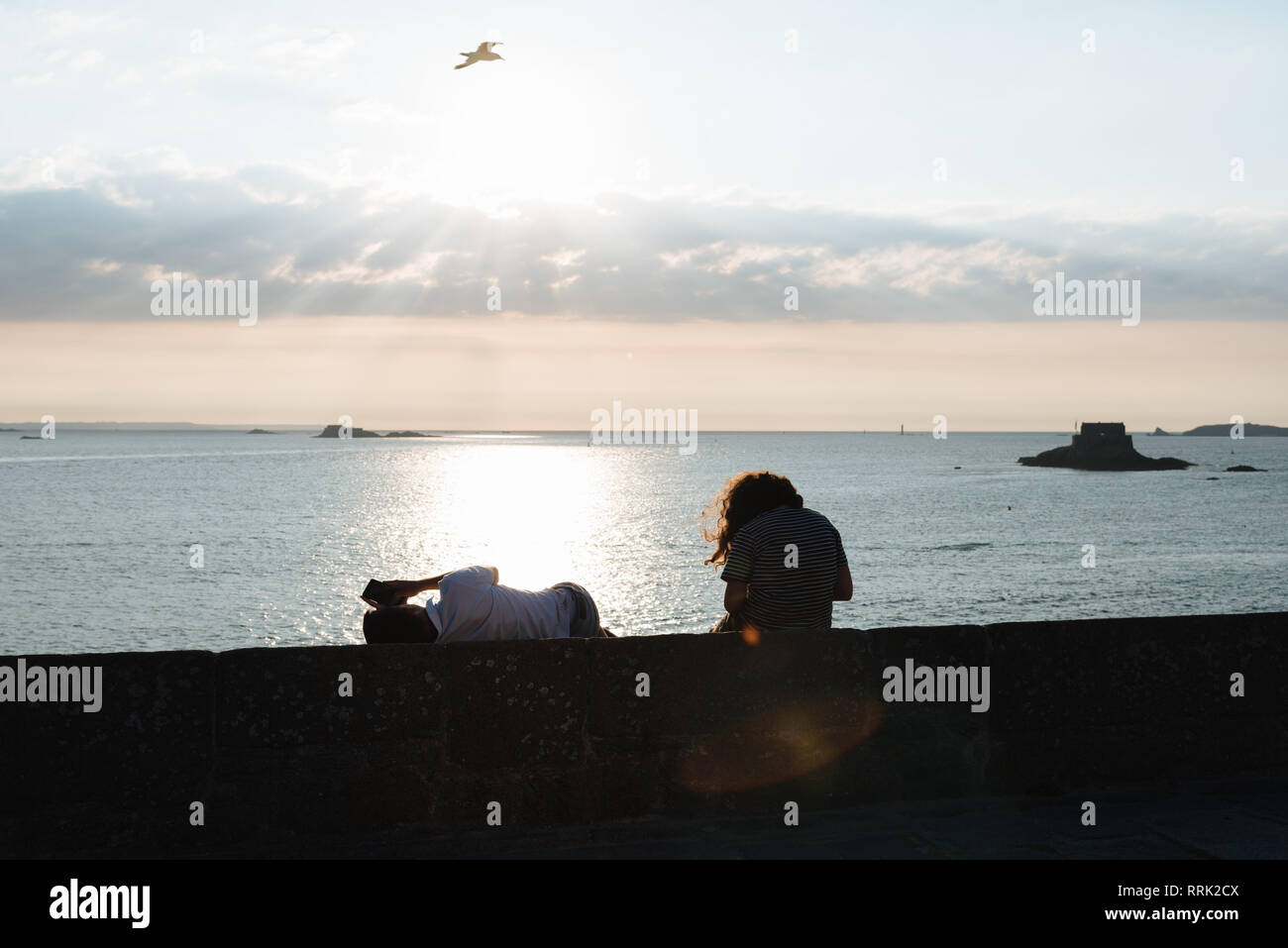 Rear view of couple fooling around while sitting on wall during sunset ...