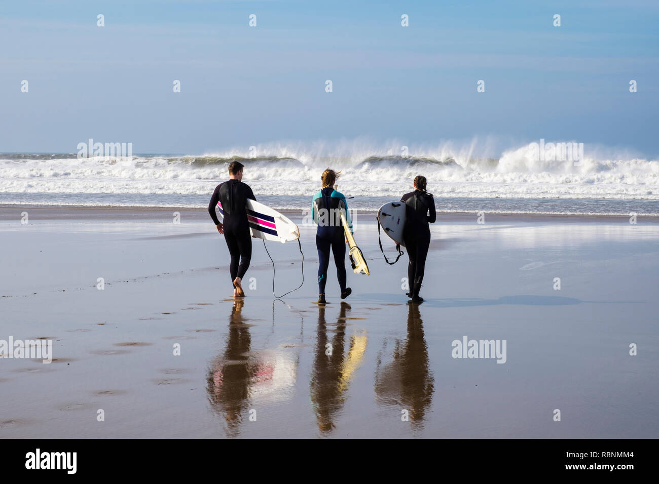 Three surfers with boards reflected in wet sand head in to the surf on ...
