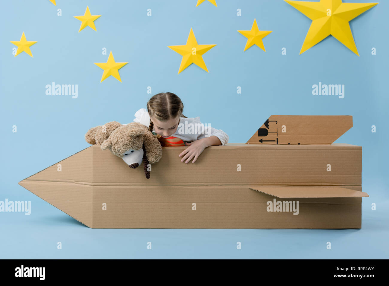 Kid with teddy bear playing with cardboard rocket on blue background ...
