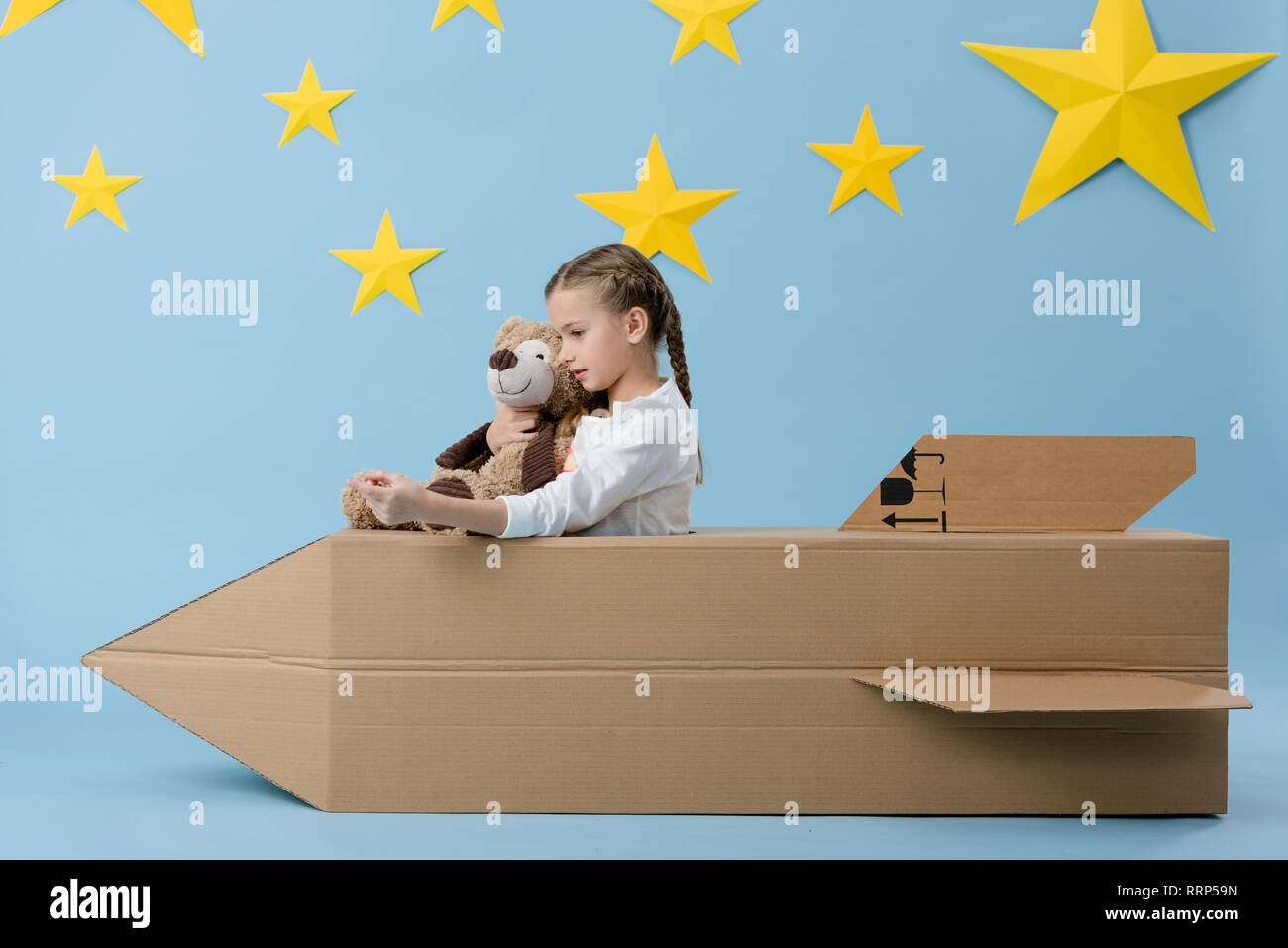 Kid holding teddy bear while playing with cardboard rocket on blue ...