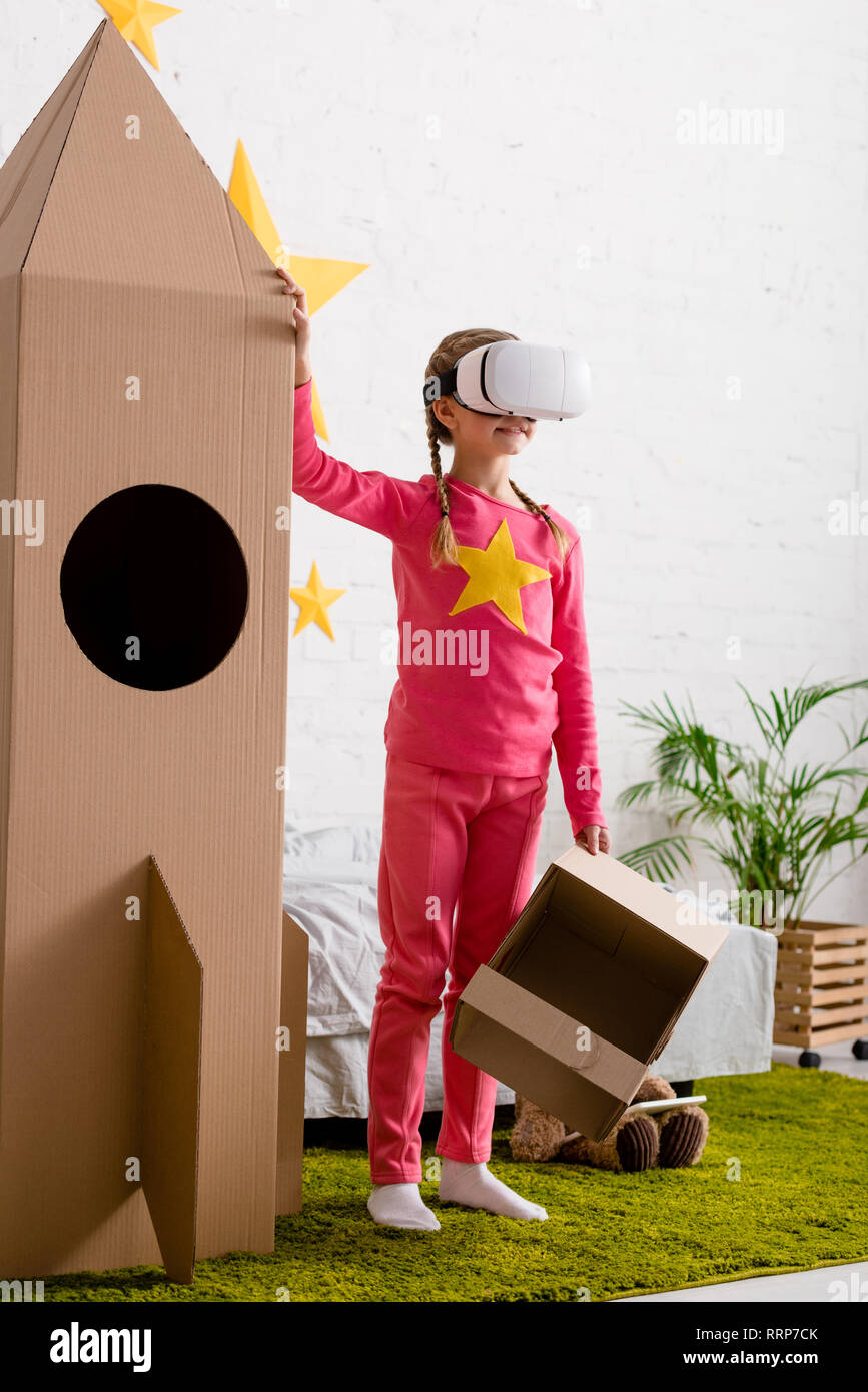 Kid in vr headset holding cardboard helmet and touching big rocket in ...