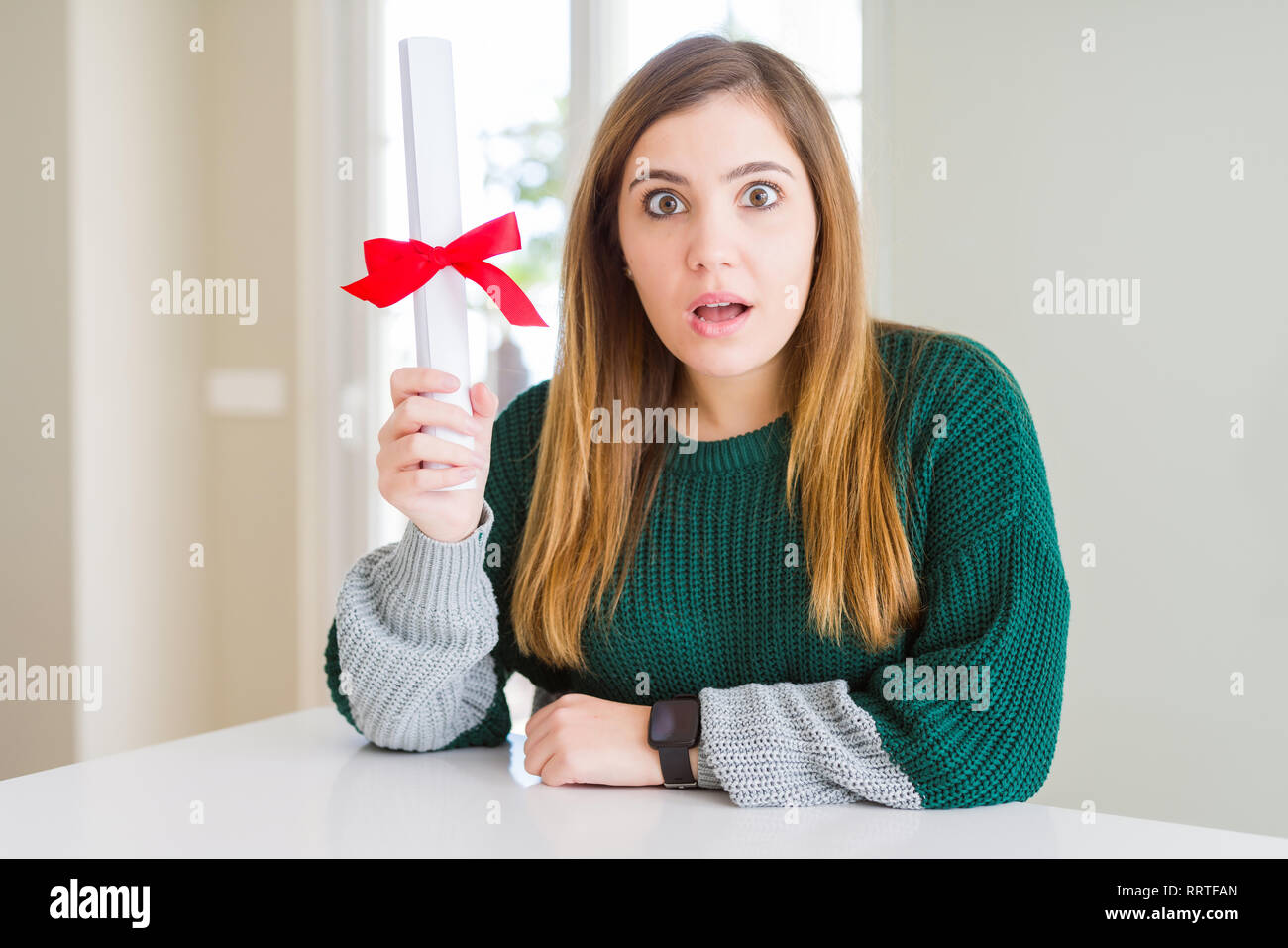 Beautiful young woman holding degree with red bow scared in shock with ...