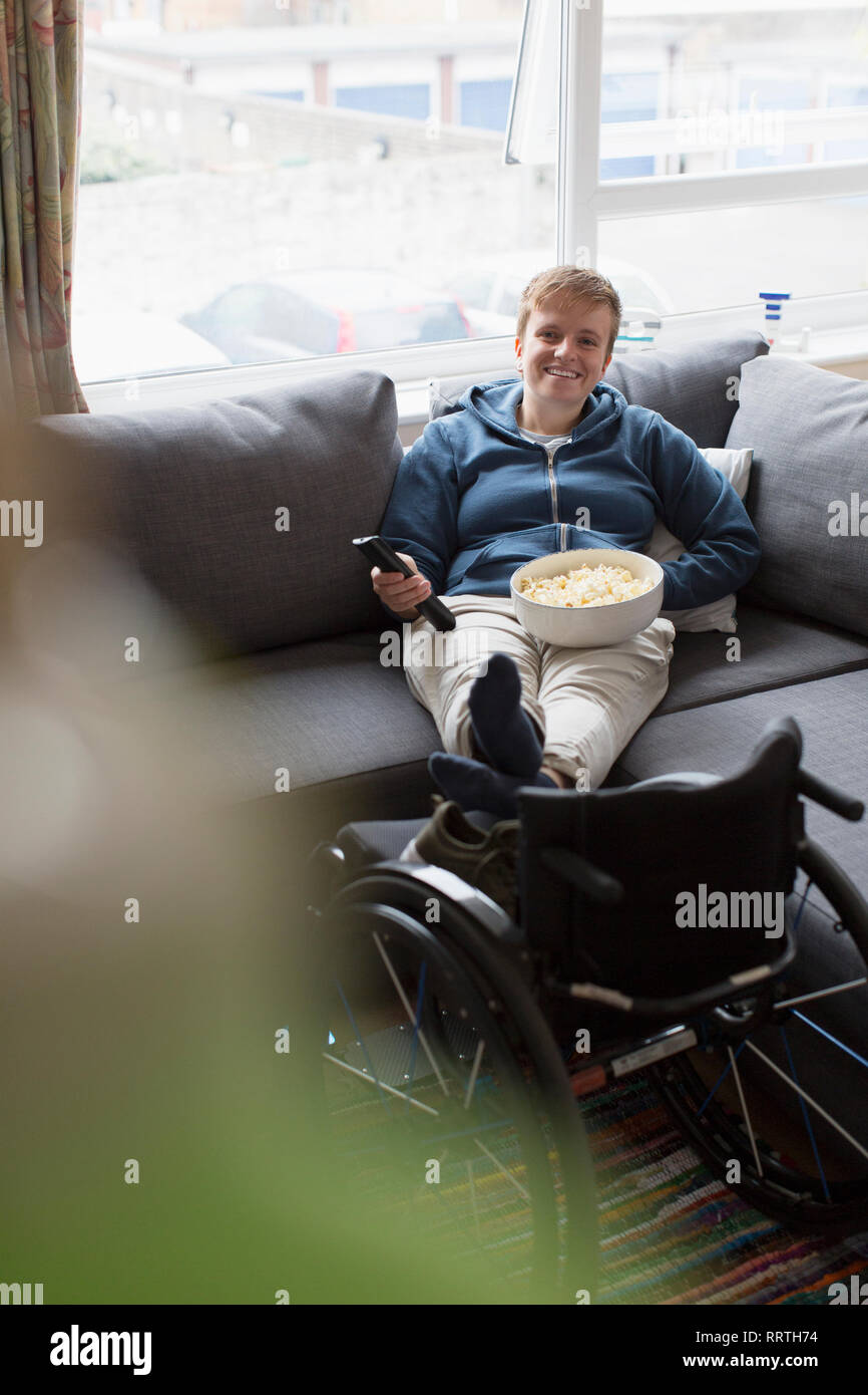 Smiling young woman watching TV and eating popcorn on sofa with feet up ...