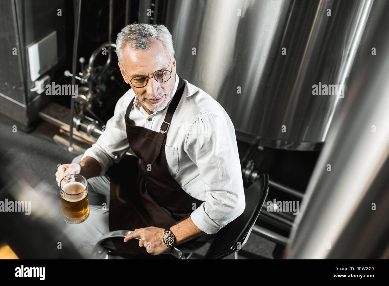 overhead view of senior brewer in apron sitting on chair and holding ...