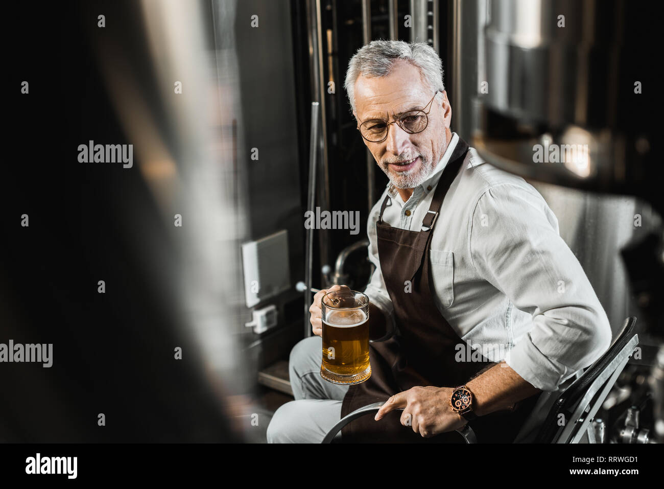 senior brewer in apron sitting on chair and holding glass of beer in ...