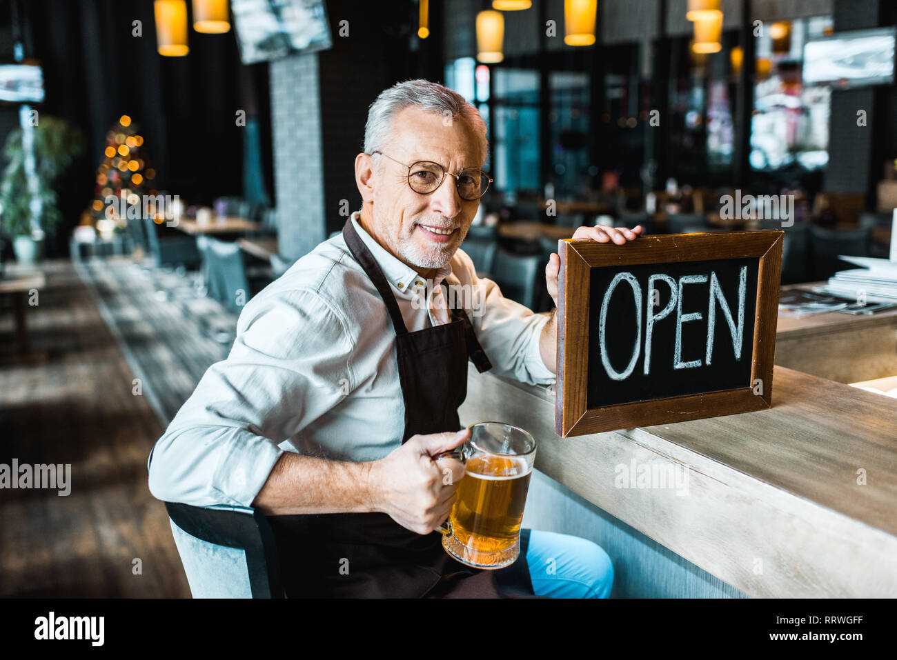 smiling owner of pub holding open sign and glass of beer at bar counter ...
