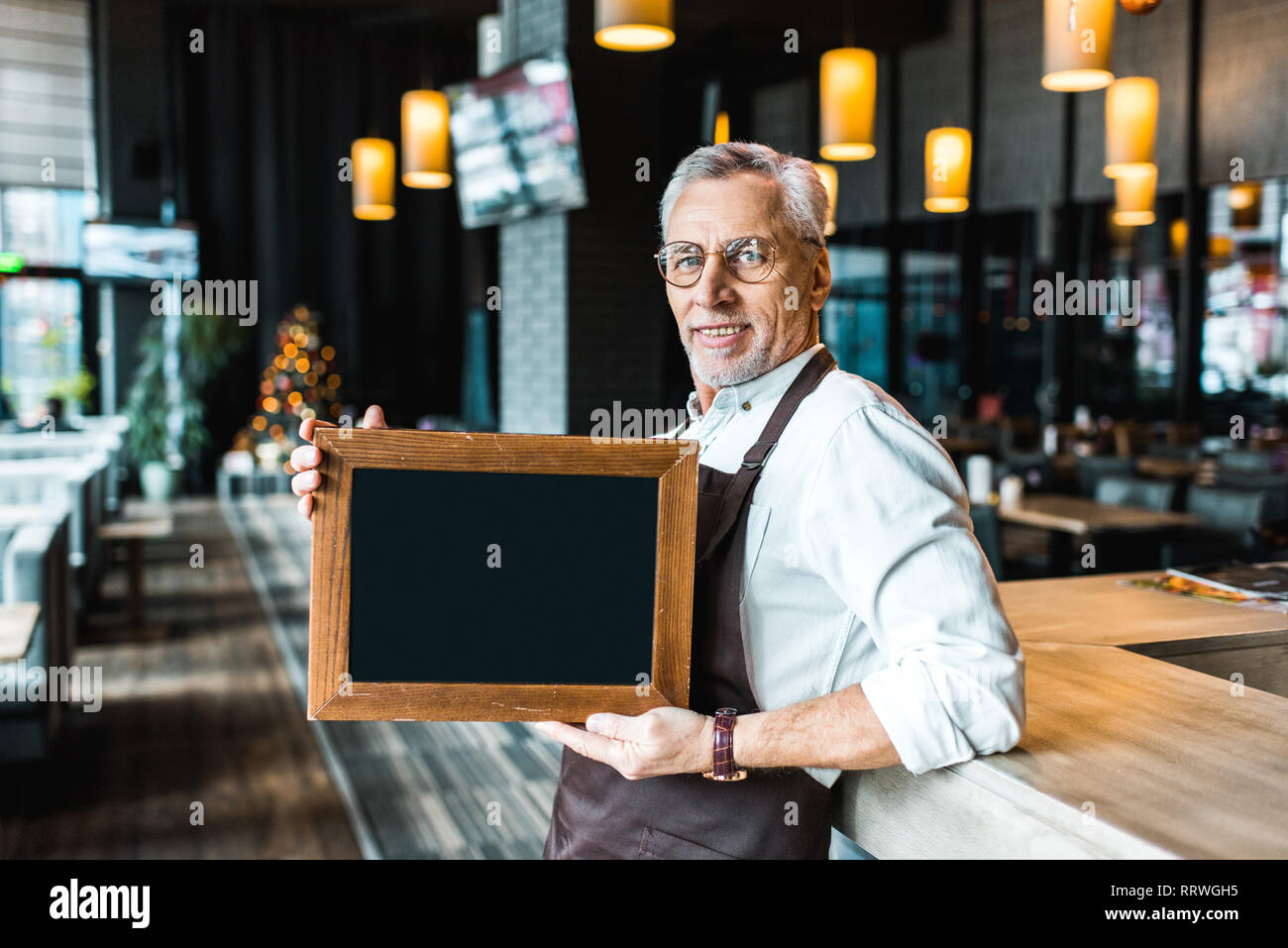 smiling owner of pub holding empty board and standing near bar counter ...