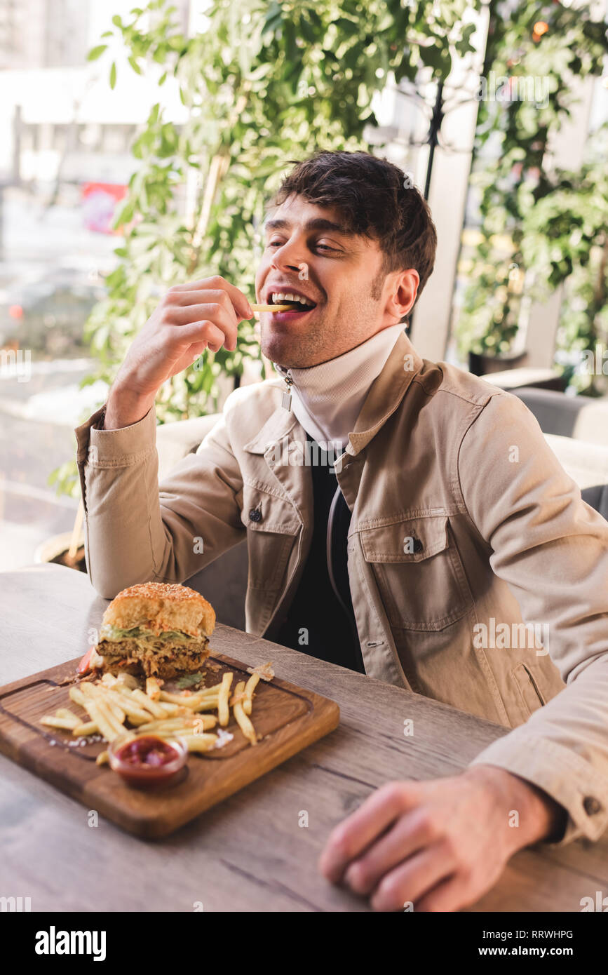 happy man eating french fry near delicious burger on cutting board in ...