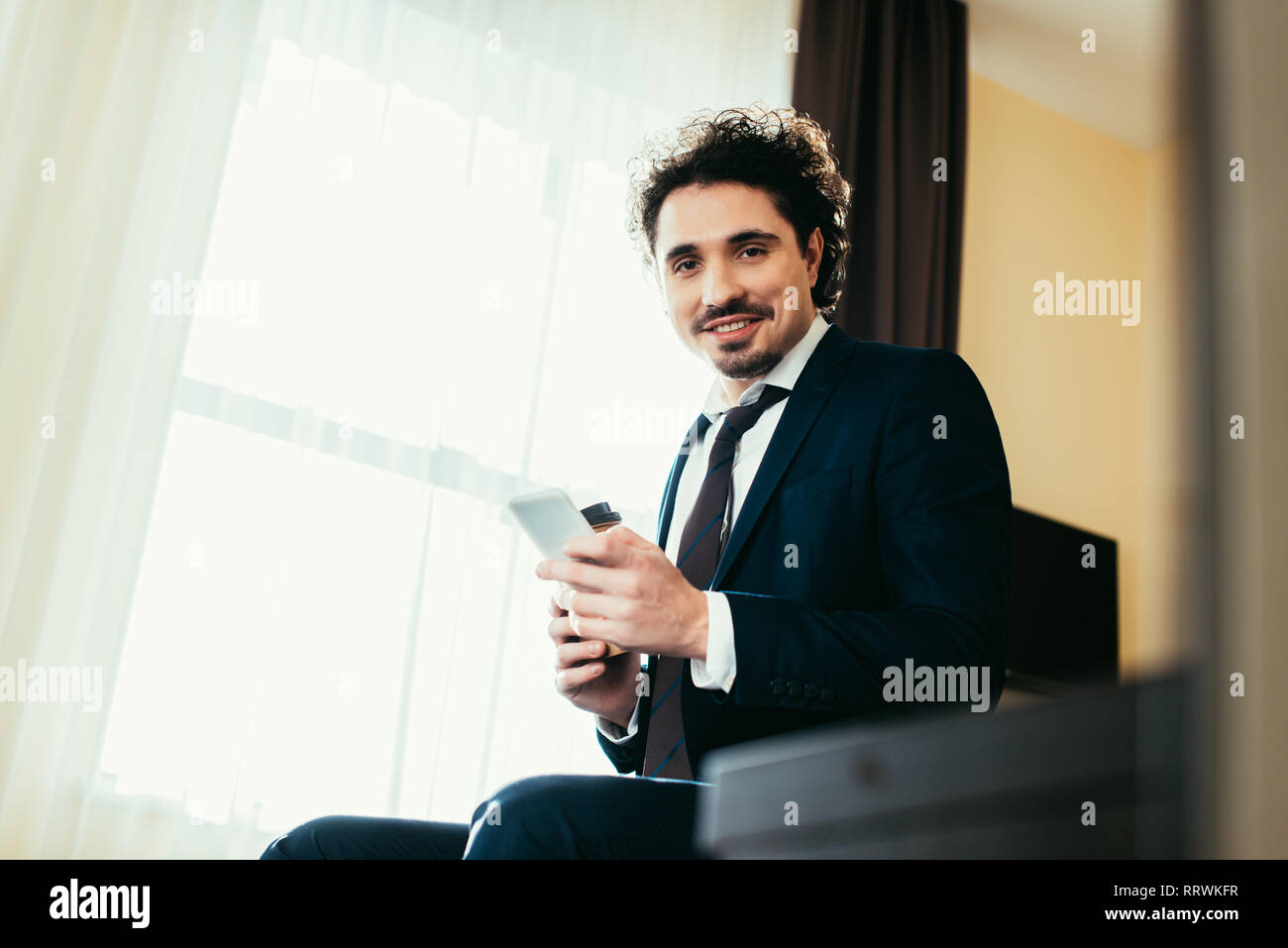 smiling businessman using smartphone during business trip in hotel room ...