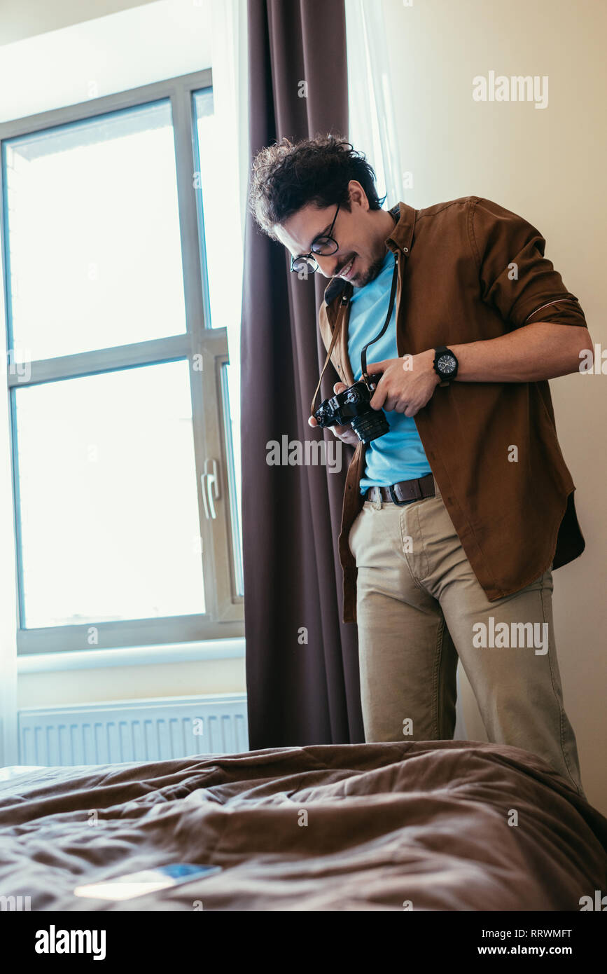 man in eyeglasses looking at photo camera near window in hotel room ...