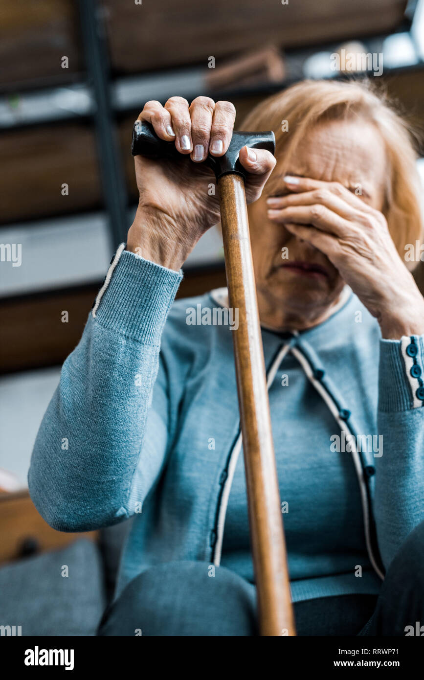 upset senior woman holding walking stick, covering face with hand and ...