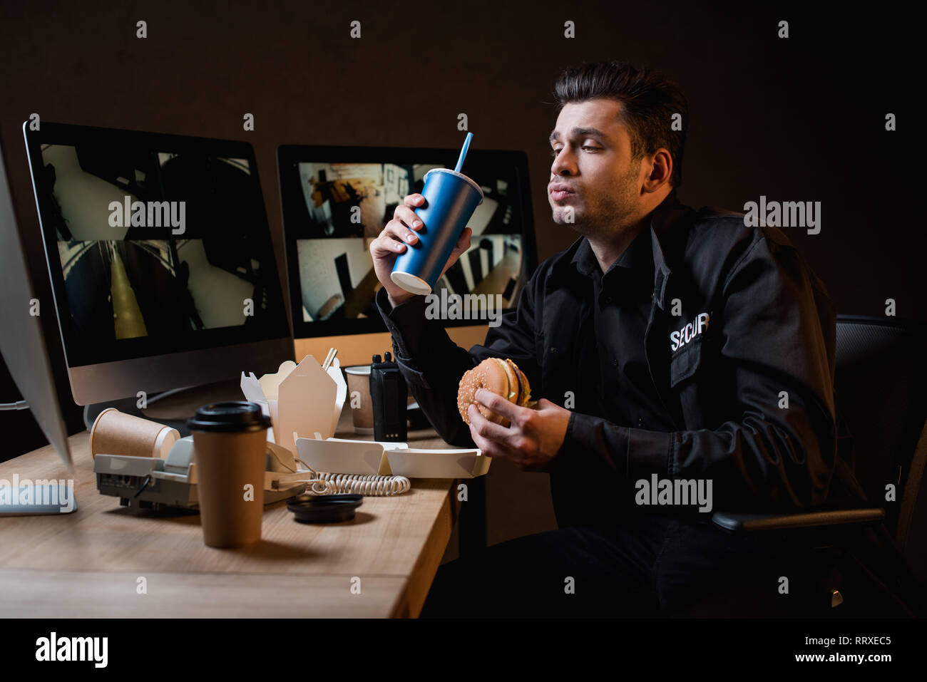 guard eating burger, holding paper cup and looking at computer monitor ...