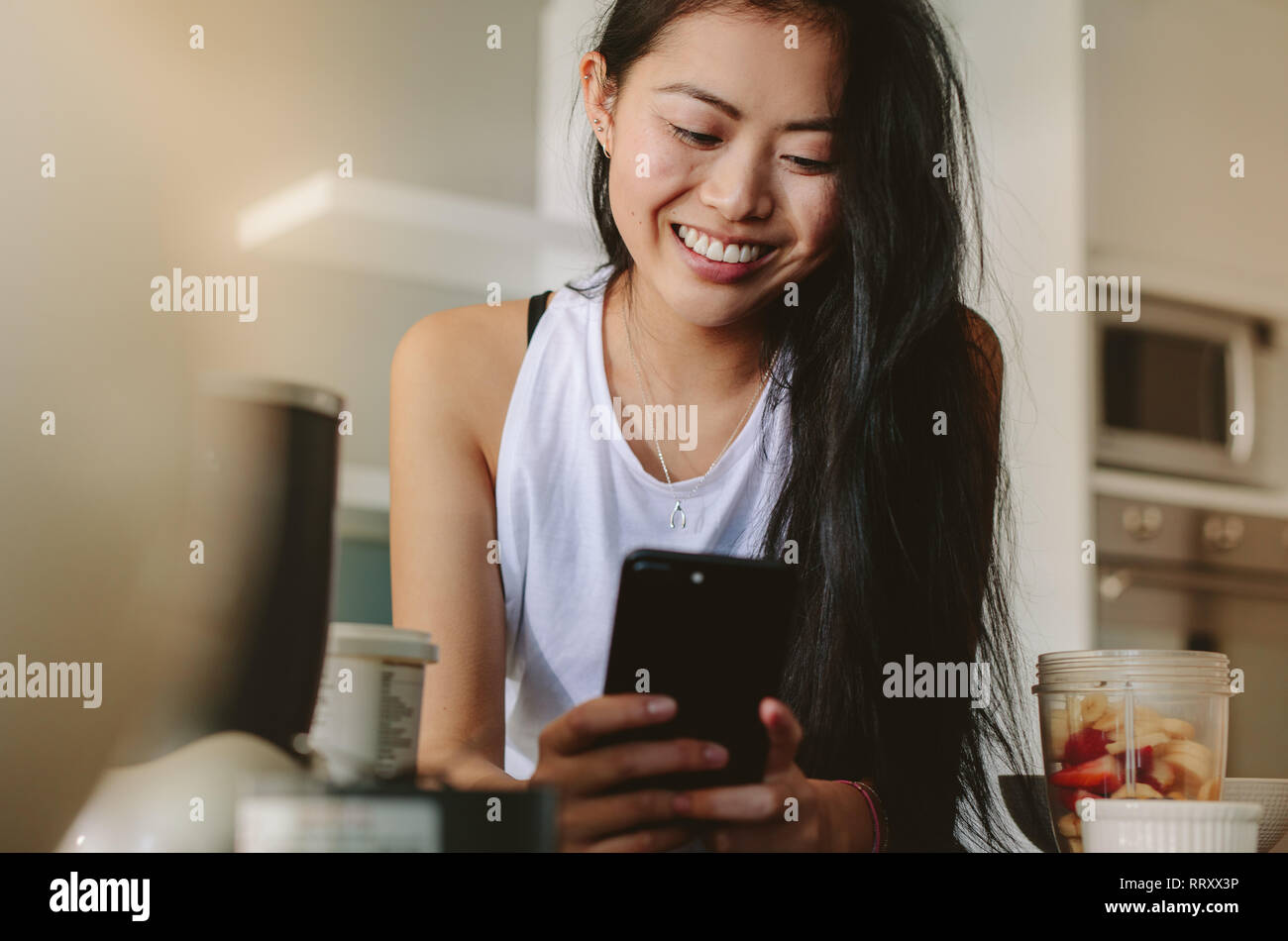 Young chinese woman using mobile phone in kitchen with fruits and ...