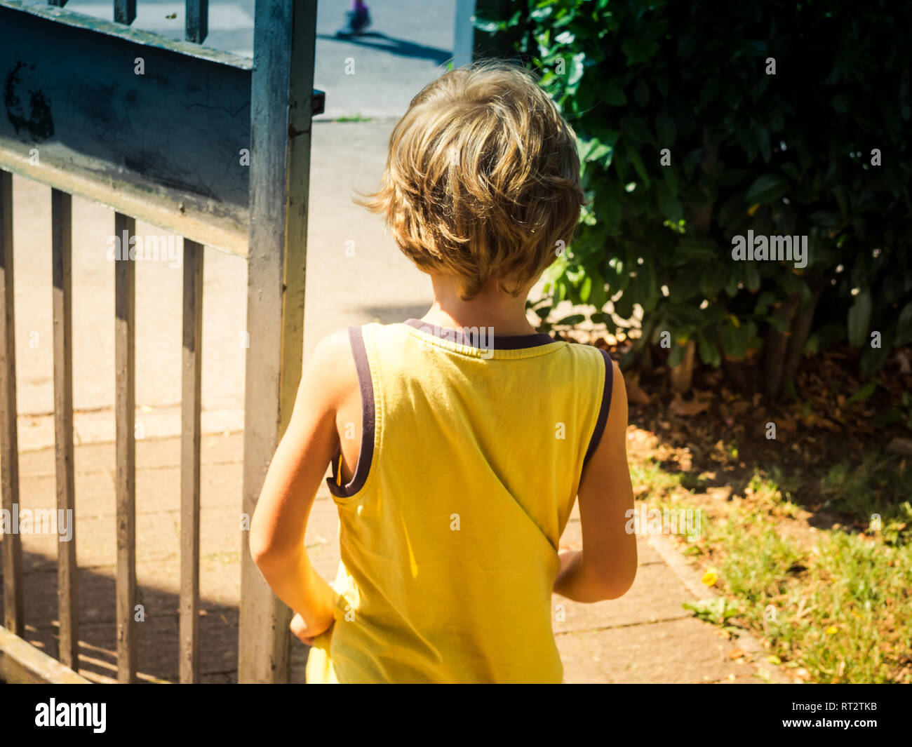 Little blonde boy on sunny day turned with his back towards the gate ...