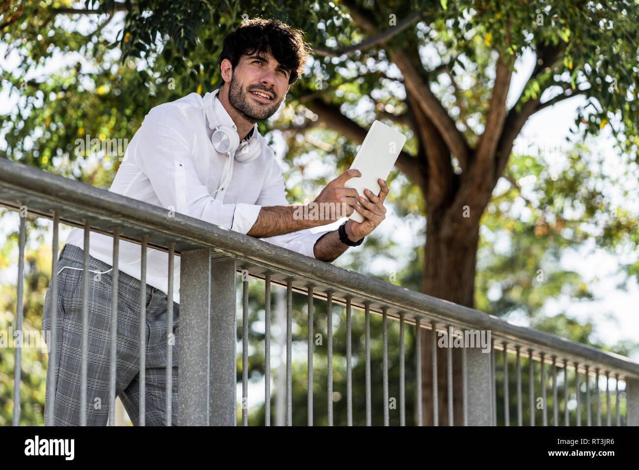 Smiling young man leaning on railing in the city holding tablet Stock ...